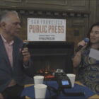 A man and a woman sitting on either side of a small table in front of a fireplace speak into handheld microphones. A sign hanging over the fireplace reads "San Francisco Public Press."