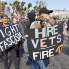 Protesters gathered outdoors carry signs, including ones with pastel letters on black poster boards reading "FIGHT FASCISM" and "HIRE VETS, FIRE 47."